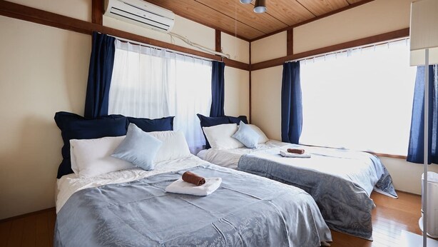 Quarto de hotel com duas camas, vigas de madeira no teto, ar condicionado e janelas amplas com cortinas azuis.