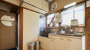 Uma cozinha compacta com armários de madeira, uma pia de aço inoxidável e uma janela sobre o balcão. Há duas lixeiras e um espelho na parede.