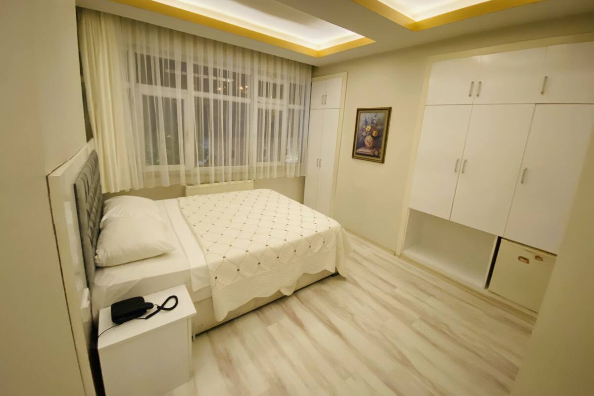 Quarto com cama, mesa de cabeceira, guarda-roupa e janela com cortinas leves.