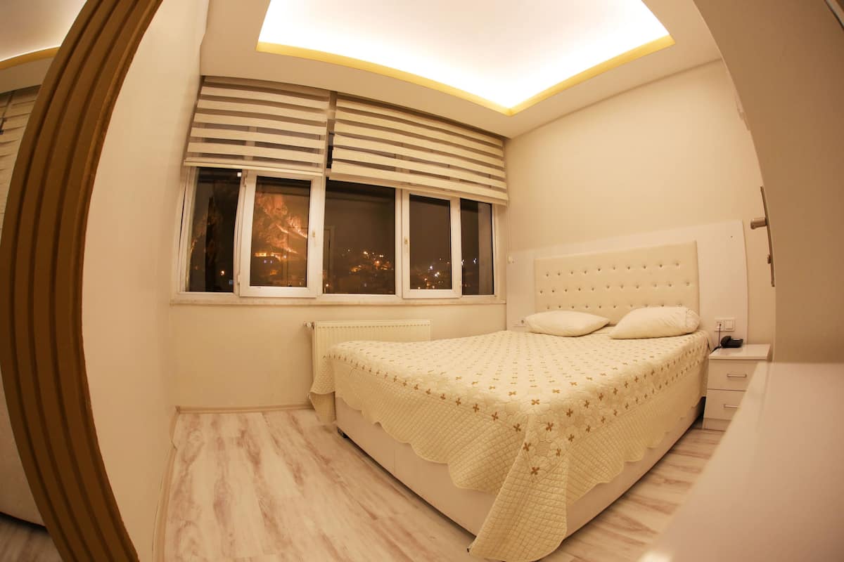 Quarto com cama, mesa de cabeceira, janela com persianas e luminária no teto.