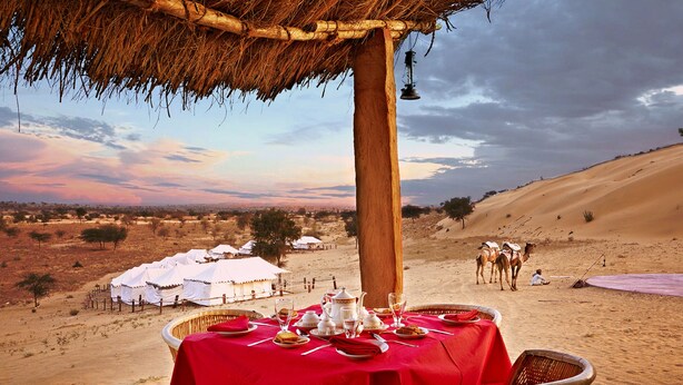 Mesa de jantar posta para dois, com toalha vermelha, rodeada por cadeiras de vime, com vista para uma paisagem desértica com tendas e camelos ao fundo.