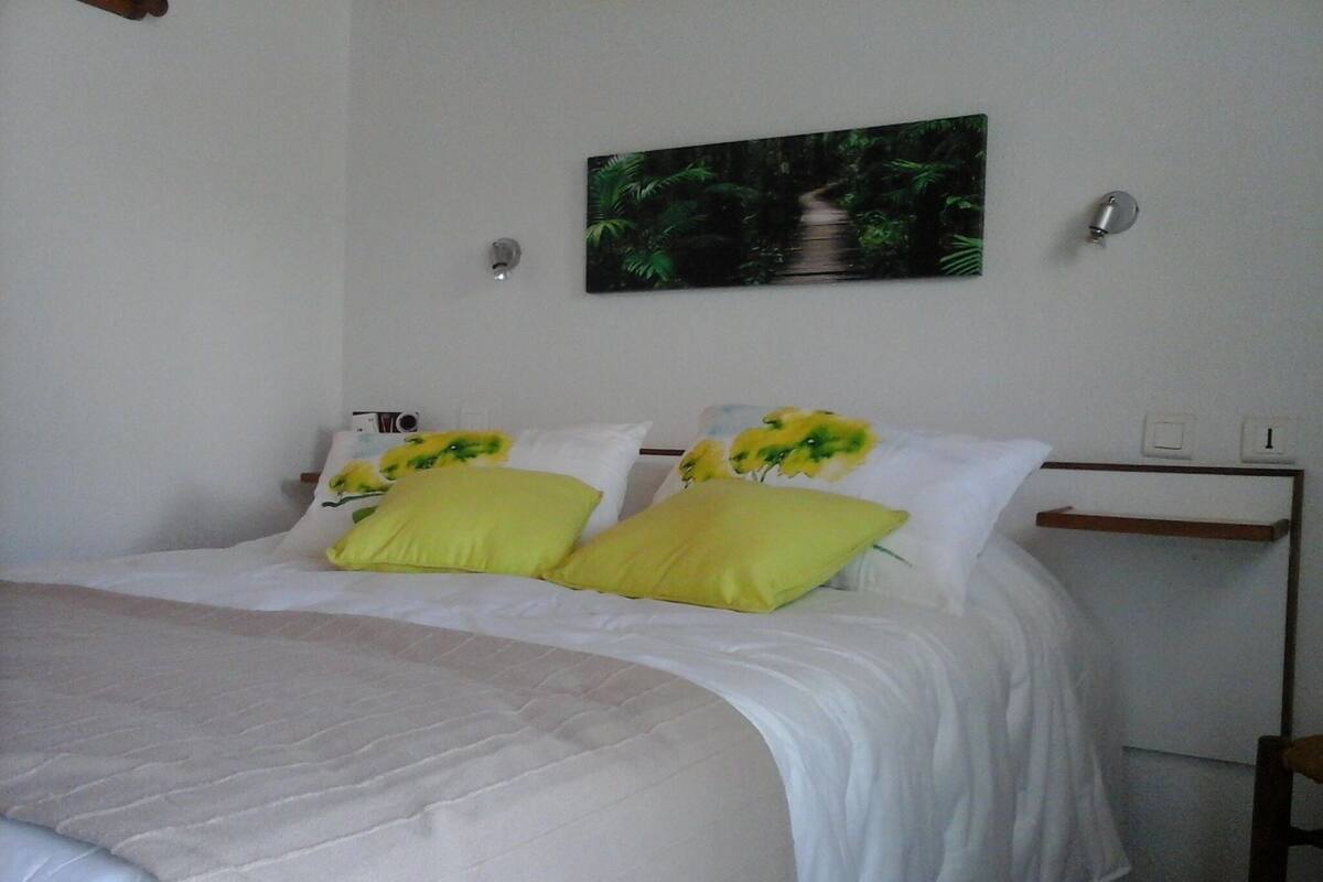 Cama bem arrumada com roupa de cama branca e duas almofadas amarelas, uma mesa de cabeceira pequena com abajur e um quadro de um caminho em meio à floresta na parede.