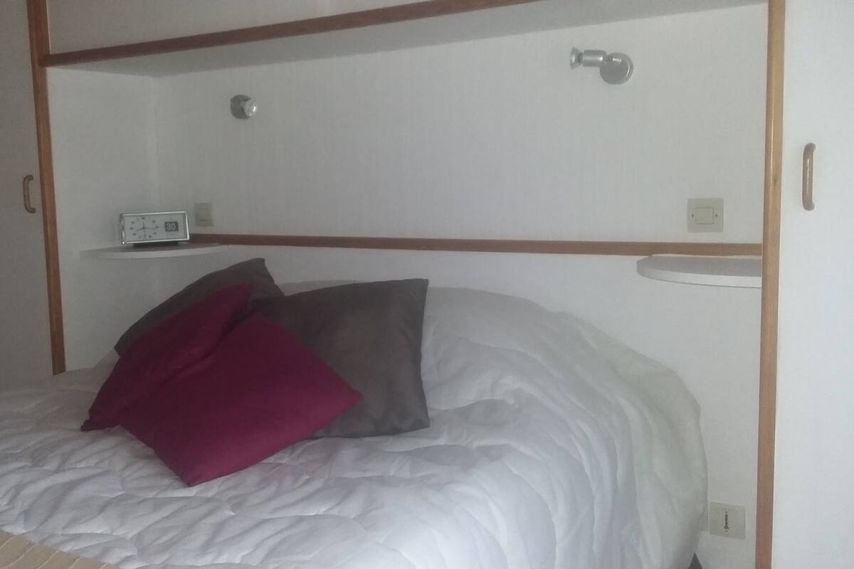 Uma cama de solteiro com colchão branco e duas almofadas, uma vermelha e uma cinza, num quarto com painéis de madeira e uma pequena mesa de cabeceira com um relógio.