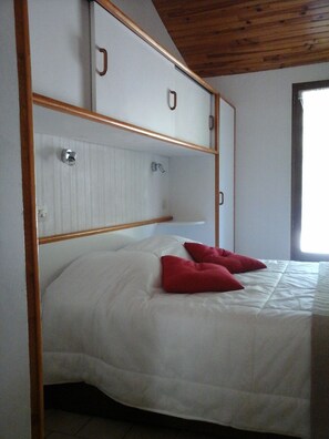 Cama com roupa de cama branca e almofadas vermelhas, cabeceira de madeira e janela com luz natural.