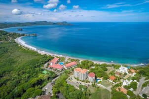 Vista aérea de um resort costeiro com villas na praia, uma piscina e vegetação exuberante.