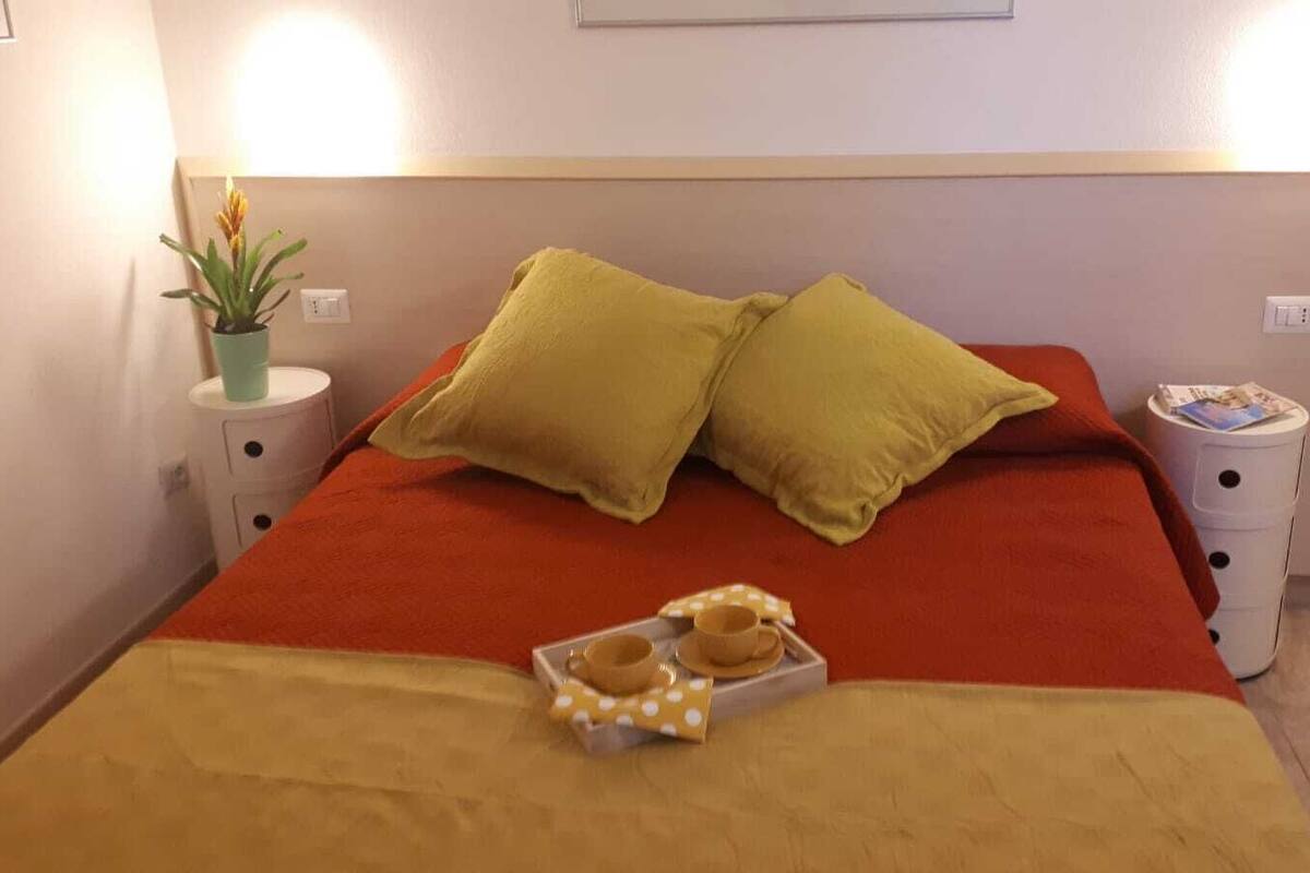 Uma cama bem arrumada com um cobertor vermelho e duas almofadas, uma mesinha de cabeceira com uma planta e um tabuleiro com chávenas e um bule.