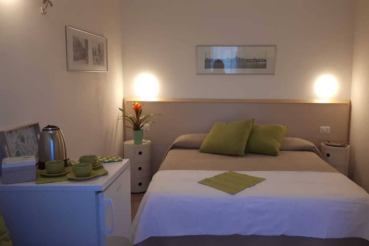 Quarto de hotel com cama, mesas de cabeceira, escrivaninha com chaleira e quadro pendurado na parede.
