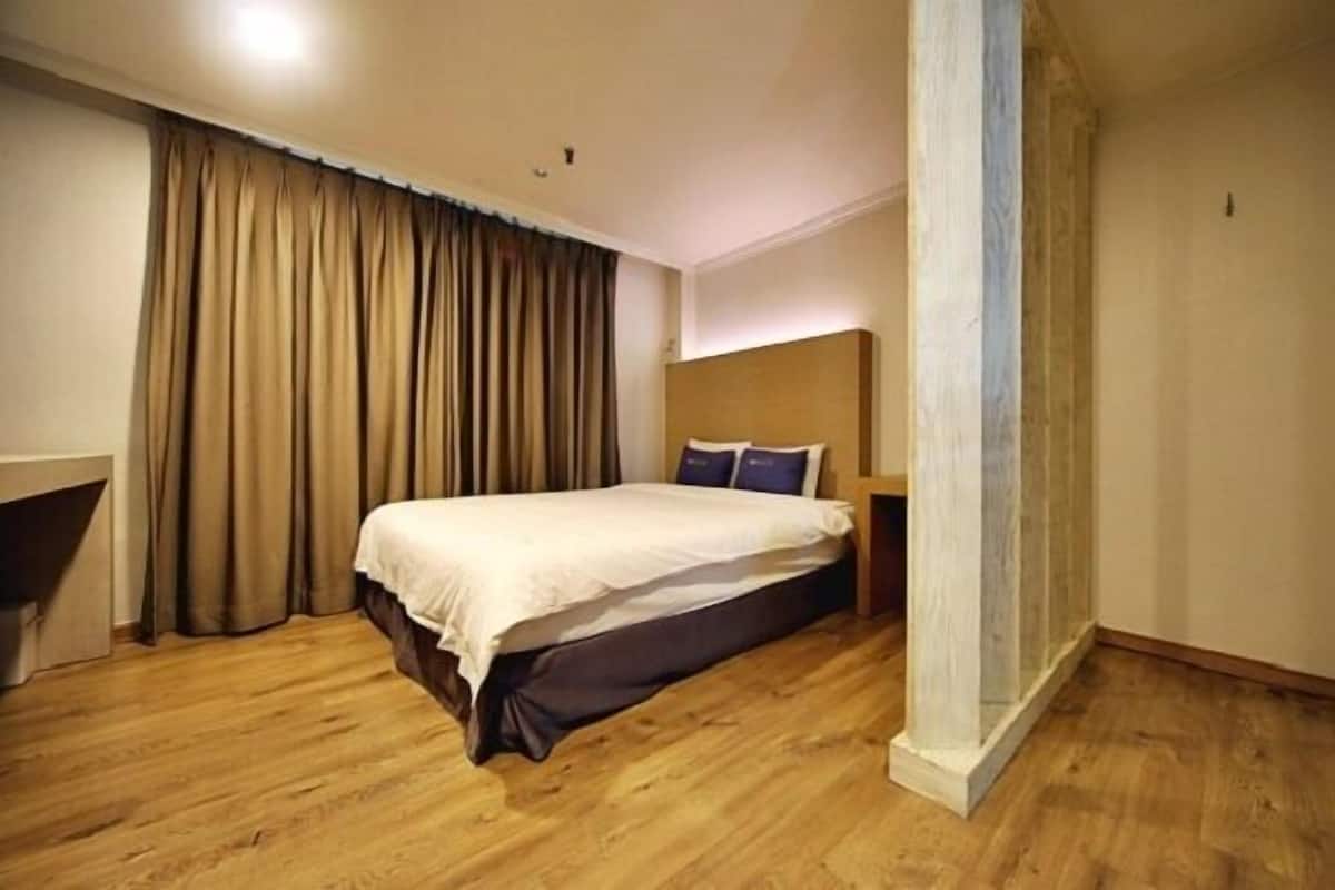 Quarto com piso de madeira, cama com roupa de cama branca, mesa de cabeceira e uma janela grande com cortinas.