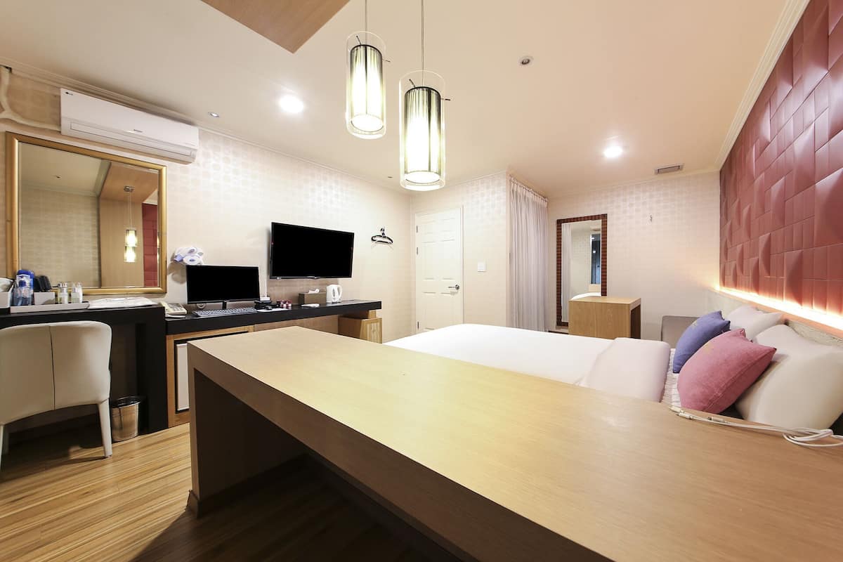 Quarto de hotel com cama, escrivaninha, dois monitores, espelho e luminárias pendentes.
