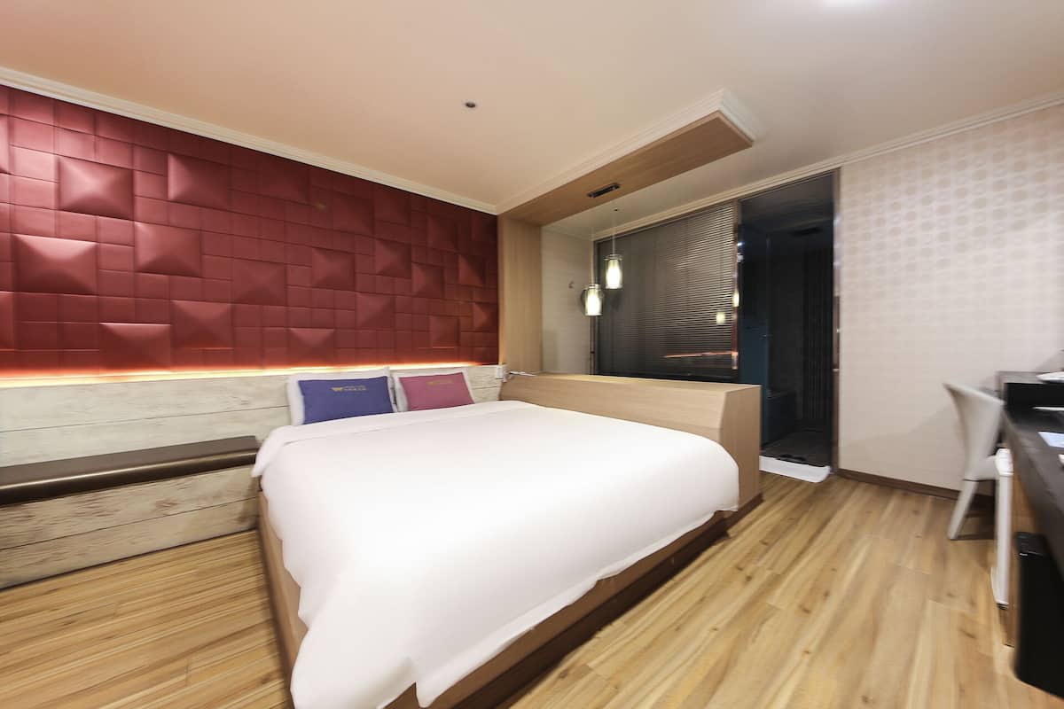 Quarto de hotel moderno com piso de madeira, cama com roupa de cama branca, cabeceira vermelha e uma janela com persianas.