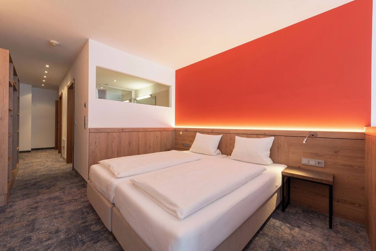 Quarto de hotel moderno com uma cama grande, uma mesa de cabeceira, um espelho e uma parede com detalhes em vermelho.