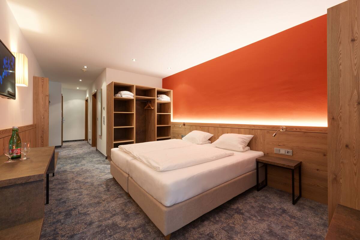 Quarto de hotel moderno com uma cama grande, guarda-roupa de madeira e uma escrivaninha com garrafa e copo.