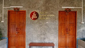 Duas portas de madeira com os números 16 e 17 em destaque acima, um banco na frente e uma parede com a inscrição "Ratti Kanchana Resort".