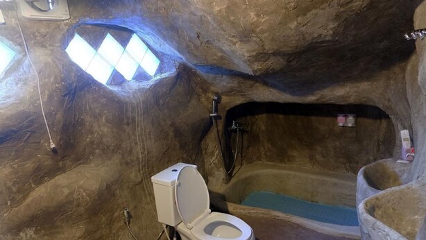 Um banheiro com parede de pedra, vaso sanitário branco, banheira e uma pequena janela.