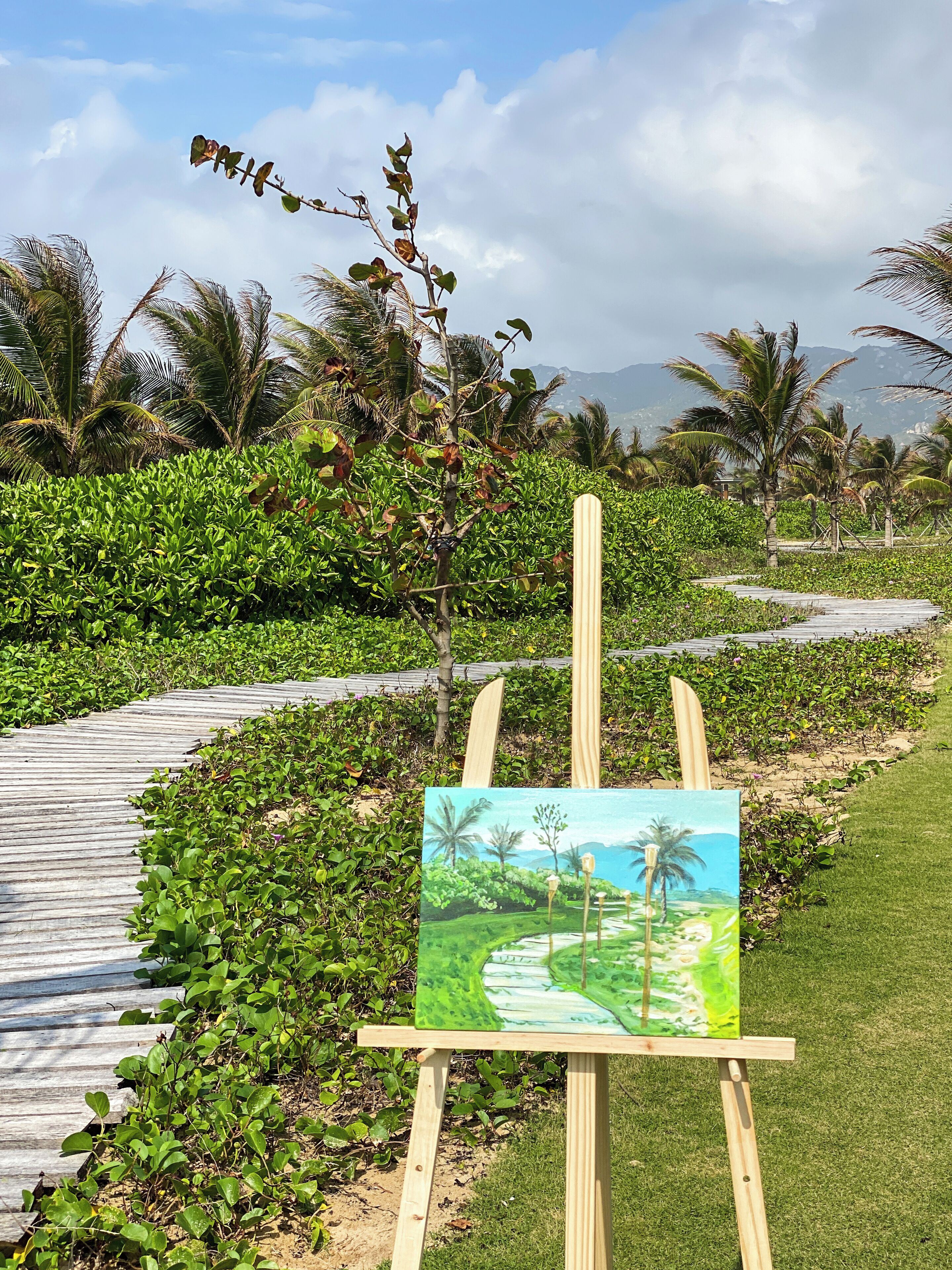 Um cavalete com uma pintura de paisagem tropical, rodeado por vegetação exuberante e palmeiras.