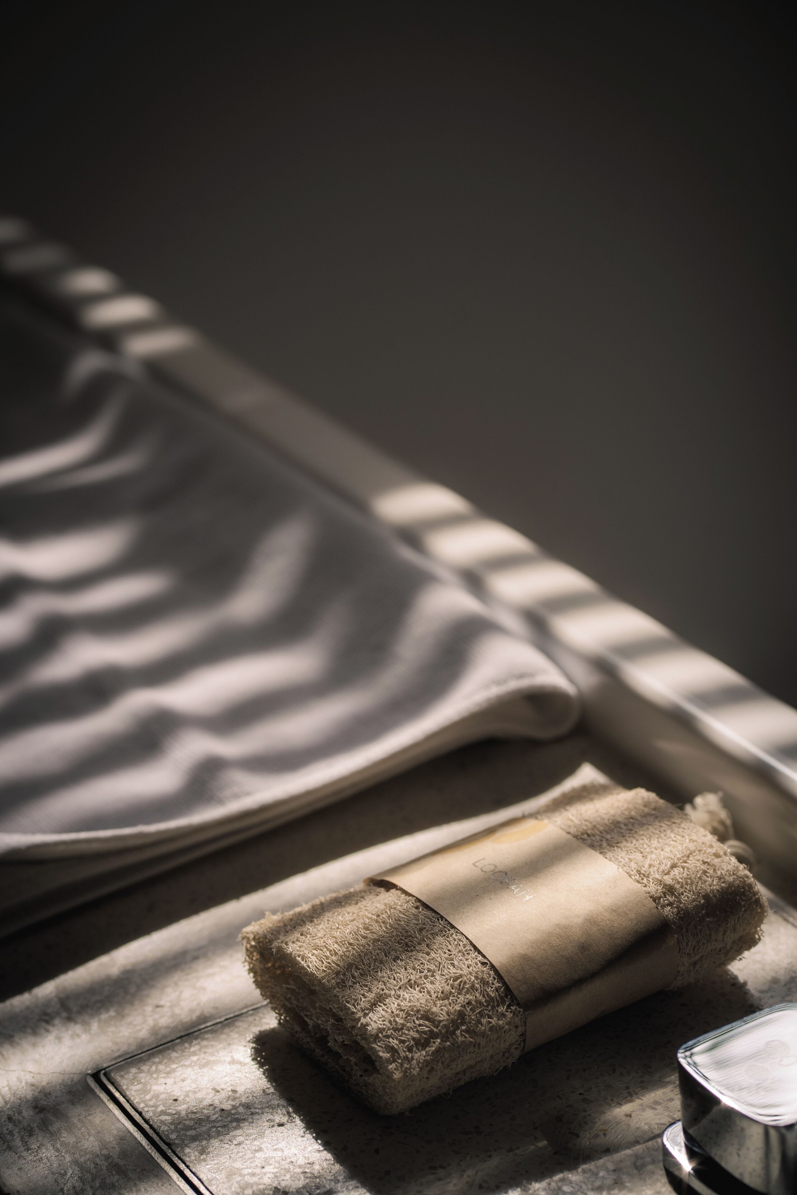 Toalha bem dobrada sobre uma cama, com etiqueta visível, um recipiente de metal e almofada estampada.