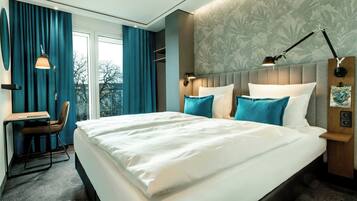 Quarto de hotel com uma cama grande, uma escrivaninha e uma cadeira. Há uma janela com cortinas, uma parede com padrão floral e uma mesa de cabeceira com abajur.
