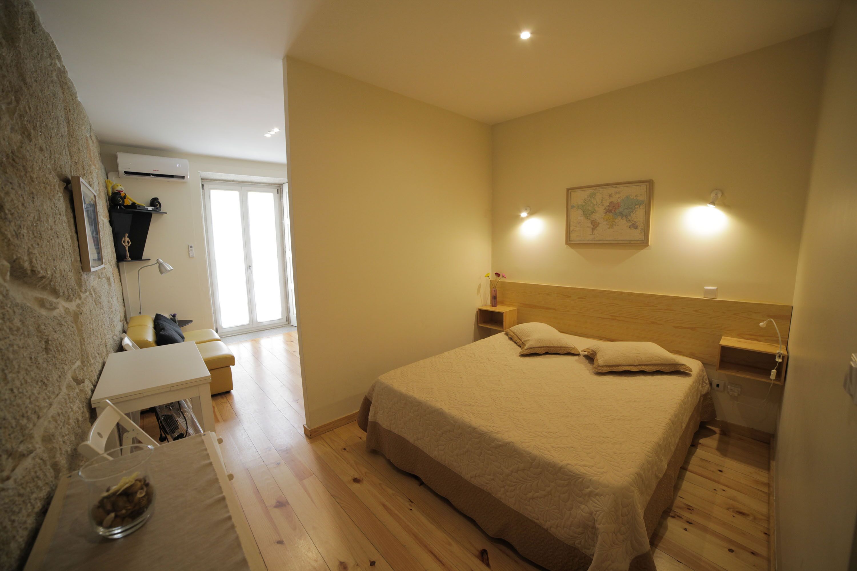 Quarto com cama grande, mesinha, cadeira, prateleira com objetos e um mapa na parede.