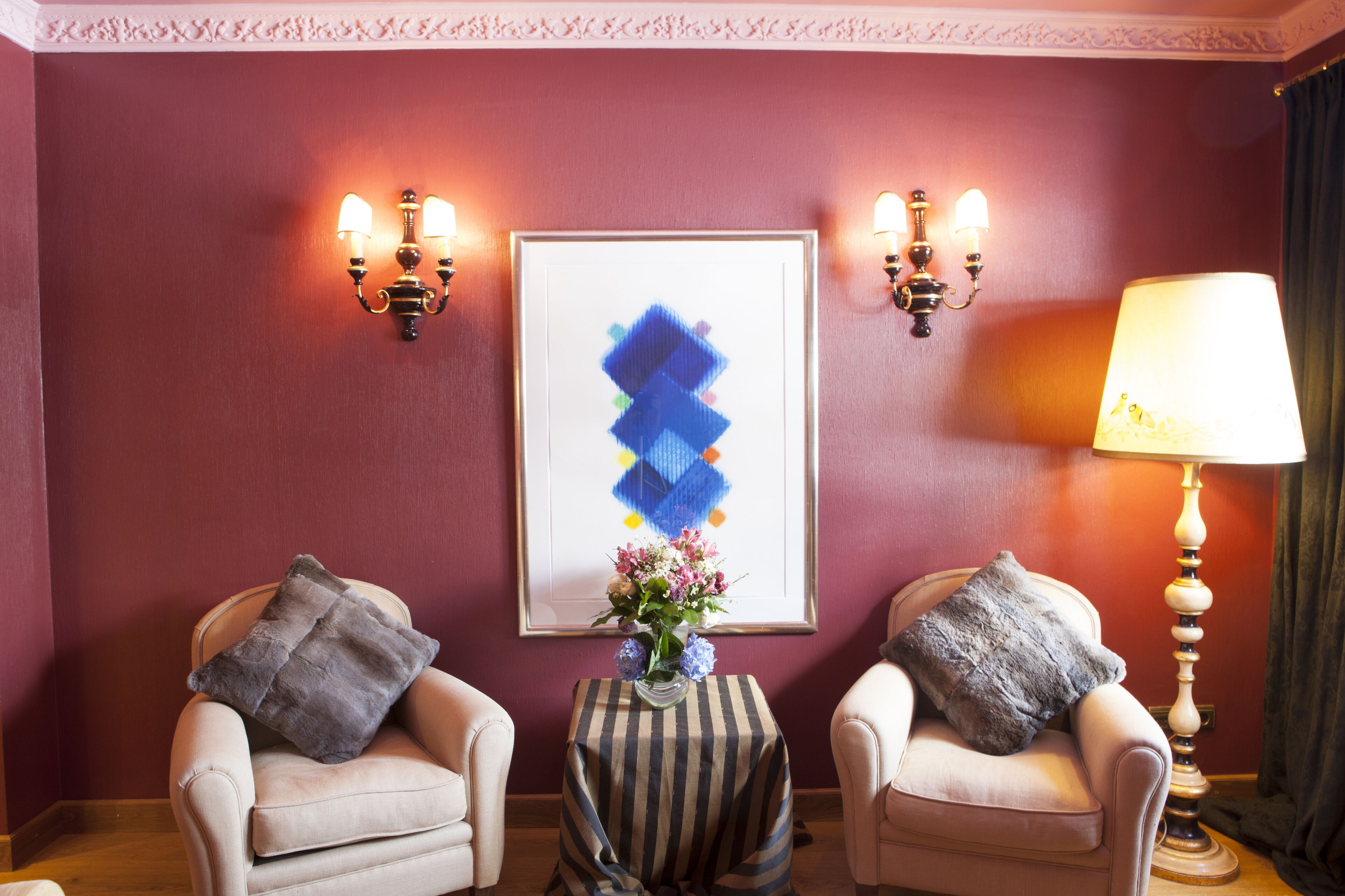 Quarto com duas poltronas, uma mesinha com um vaso de flores, um quadro abstrato emoldurado e luminárias na parede.