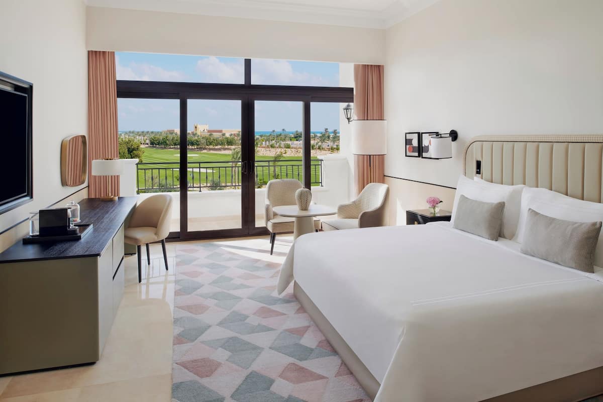 Quarto de hotel com uma cama grande, uma escrivaninha, uma cadeira, uma mesa pequena e uma varanda com vista para um campo de golfe.
