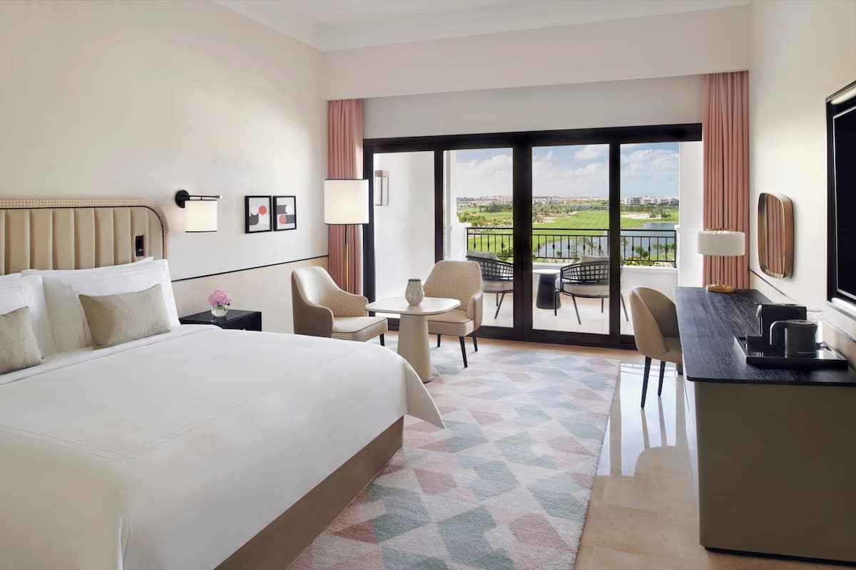 Quarto de hotel com uma cama grande, uma escrivaninha, uma cadeira, uma mesa pequena e uma varanda com vista para a cidade.