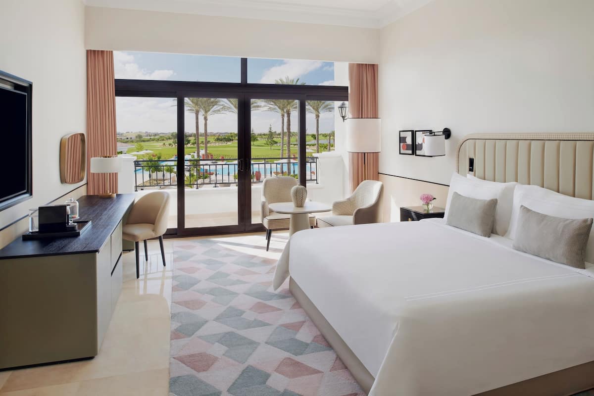 Quarto de hotel com uma cama grande, uma escrivaninha com televisão, uma mesinha e cadeiras. O quarto tem vista para uma piscina e palmeiras.
