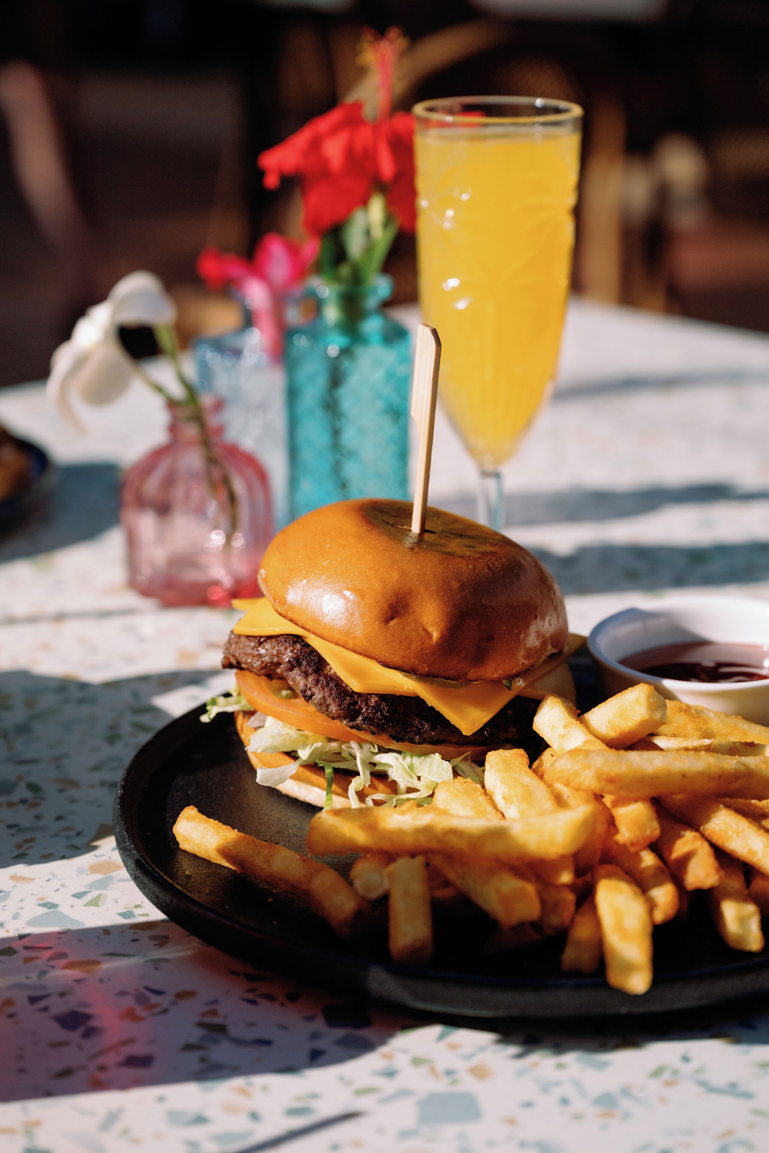 Um hambúrguer com batatas fritas em um prato preto, um copo de suco de laranja e um vaso com flores ao fundo.