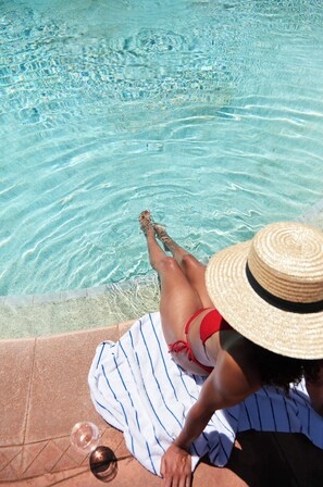 Uma pessoa relaxando à beira de uma piscina com água cristalina, usando chapéu de palha e roupas listradas.