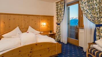 Uma cama de madeira com roupa de cama branca, uma mesa de cabeceira com abajur e vista para uma montanha através de uma janela com cortinas florais.
