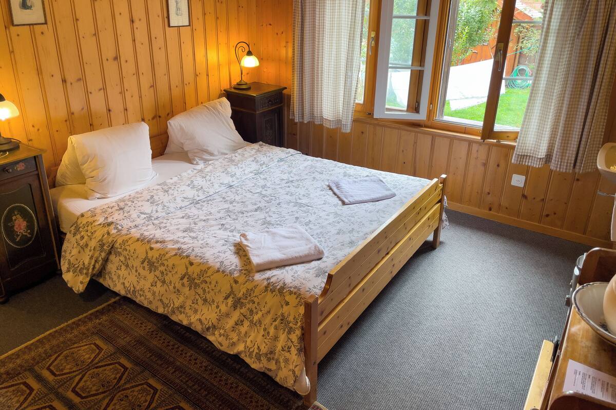 Quarto com painéis de madeira, uma cama, mesa de cabeceira, abajur e janela com cortinas.