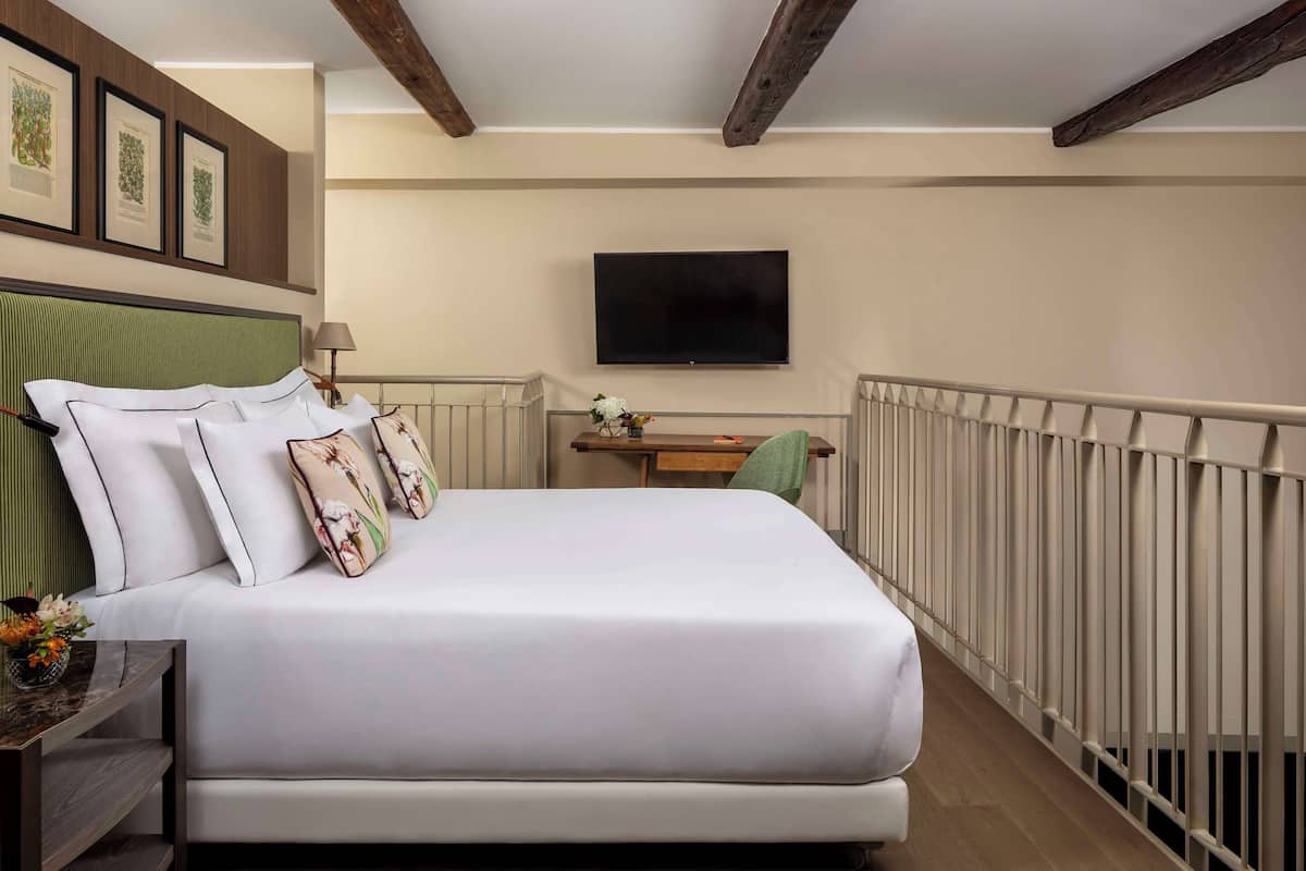Quarto de hotel com uma cama grande, televisão de tela plana na parede, escrivaninha com cadeira e corrimão junto à janela.