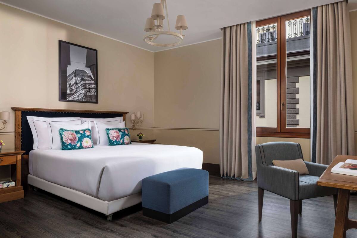 Quarto de hotel com uma cama grande, uma cadeira, uma mesa e uma janela com cortinas.