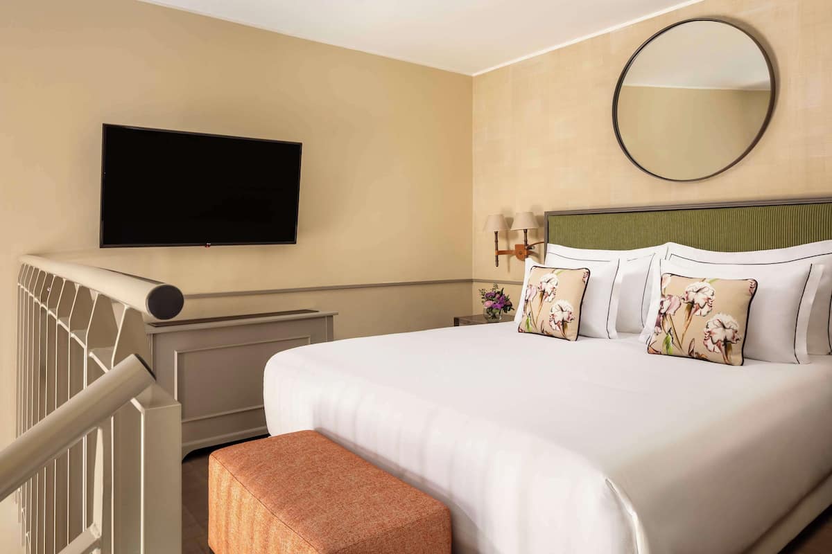 Quarto de hotel com uma cama grande, uma TV de tela plana fixada na parede, um espelho redondo e uma mesinha de cabeceira com abajur.