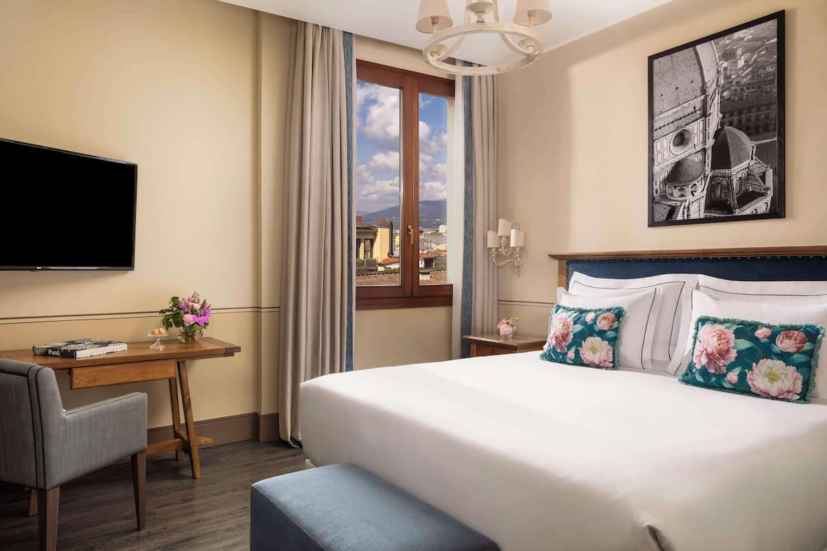Quarto de hotel com cama, escrivaninha, cadeira, televisão, janela com cortinas e vista para a cidade.