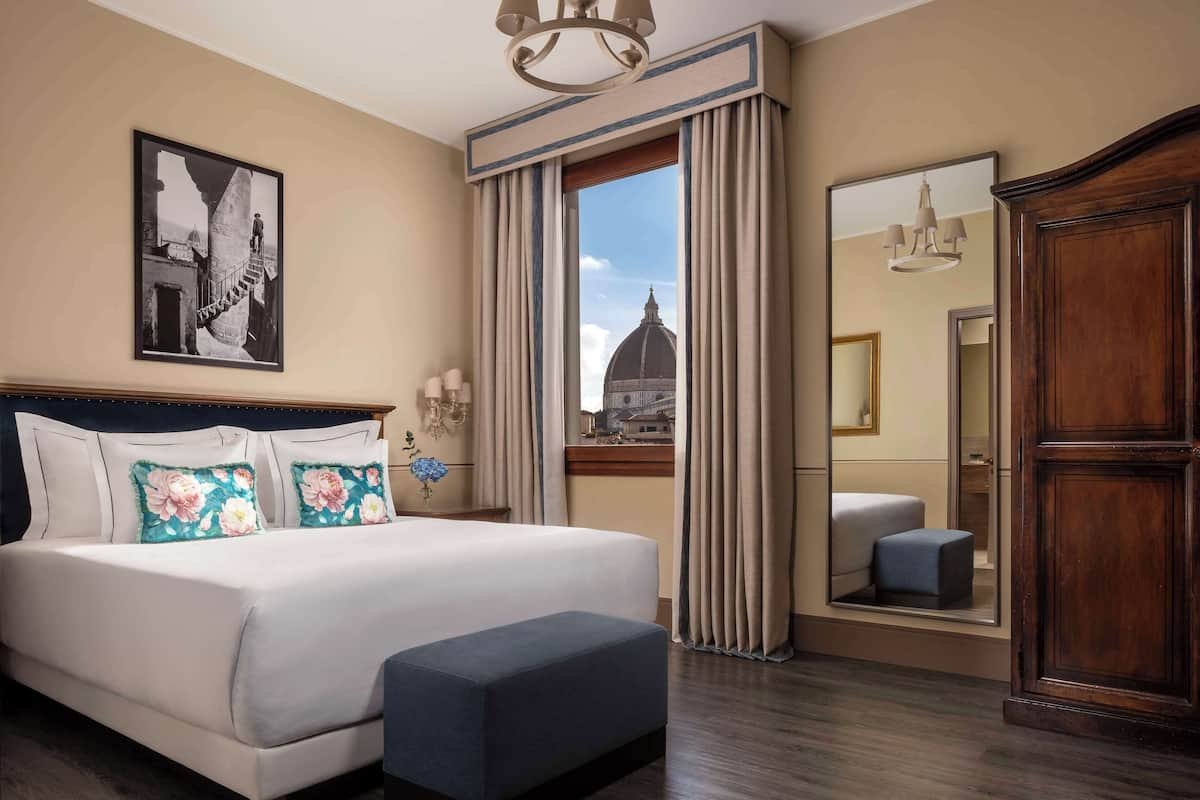 Quarto de hotel com uma cama grande, vista de uma cúpula pela janela, um espelho e um guarda-roupa de madeira.