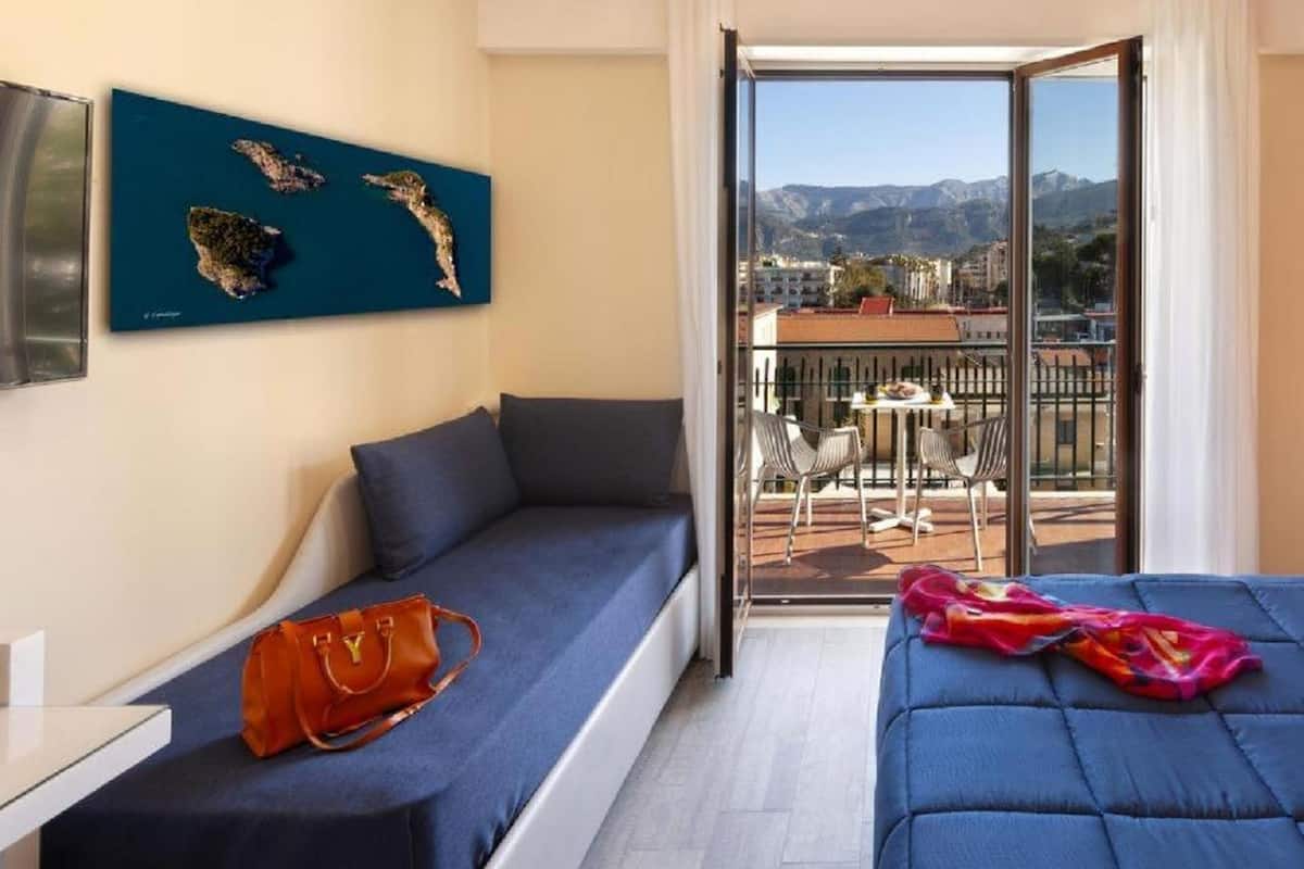 Quarto de hotel moderno com varanda, sofá, cama e vista para a cidade e montanhas.