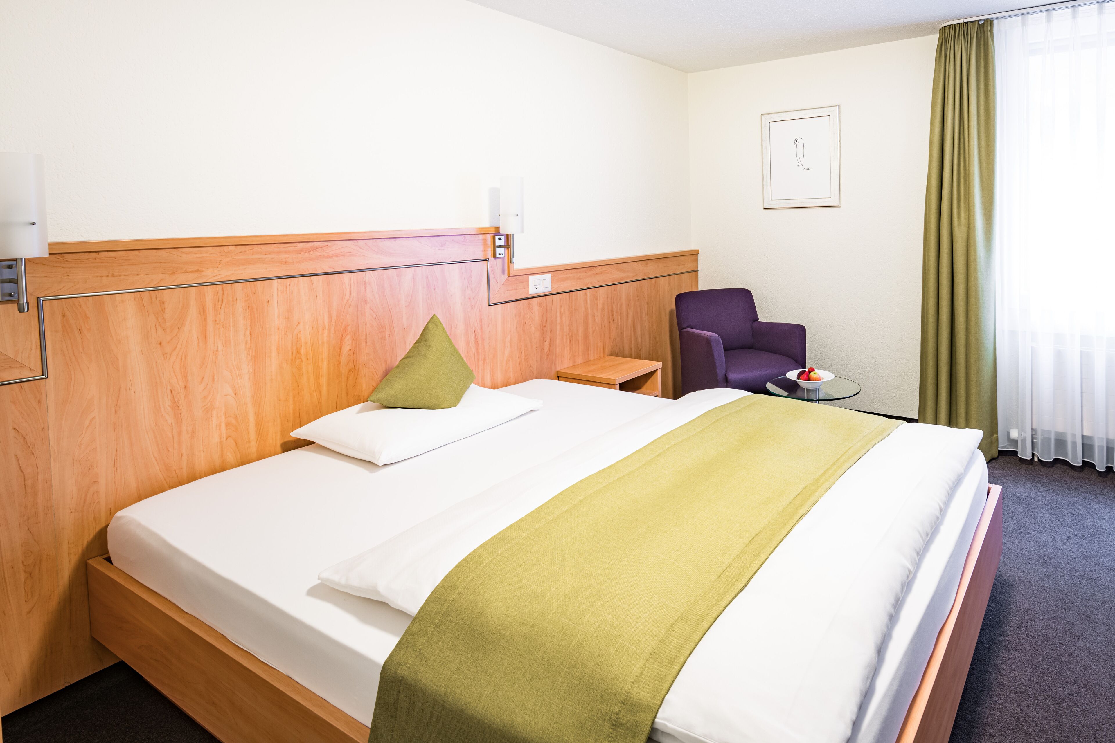 Quarto de hotel com cabeceira de madeira, cama com roupa de cama branca e um xale verde, poltrona roxa, uma pequena mesa com uma tigela de frutas e uma janela com cortina verde.