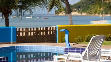 Uma área de piscina com piso de azulejos, cerca de madeira e duas cadeiras de descanso brancas.