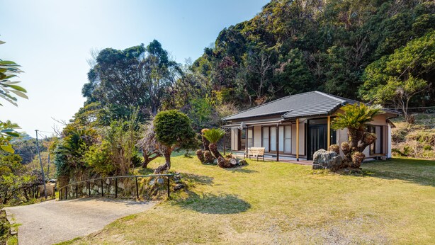 Uma casa tradicional japonesa com telhado de telhas, cercada por um jardim bem cuidado com diversas plantas e um caminho de pedras que leva à entrada.
