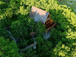 Vista aérea de uma casa cercada por vegetação verde densa, com um deck de madeira visível e um caminho de pedra que leva à entrada.