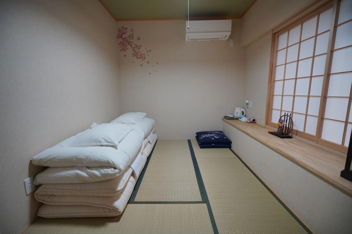 Quarto japonês tradicional com tatames, futon, uma pequena mesa com chaleira e janela com portas shoji.