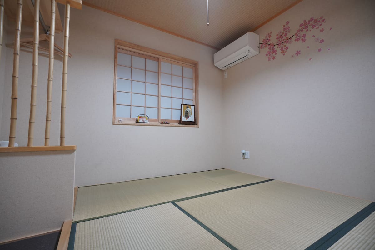 Quarto com piso de tatame, janela com tela shoji, ar-condicionado fixado na parede e um ramo decorativo de flor de cerejeira.