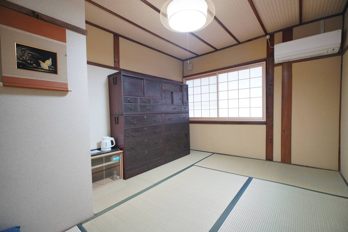 Um quarto japonês tradicional com tatame, um armário de madeira escura, uma pequena mesa com um chaleira e uma janela com padrão quadriculado.