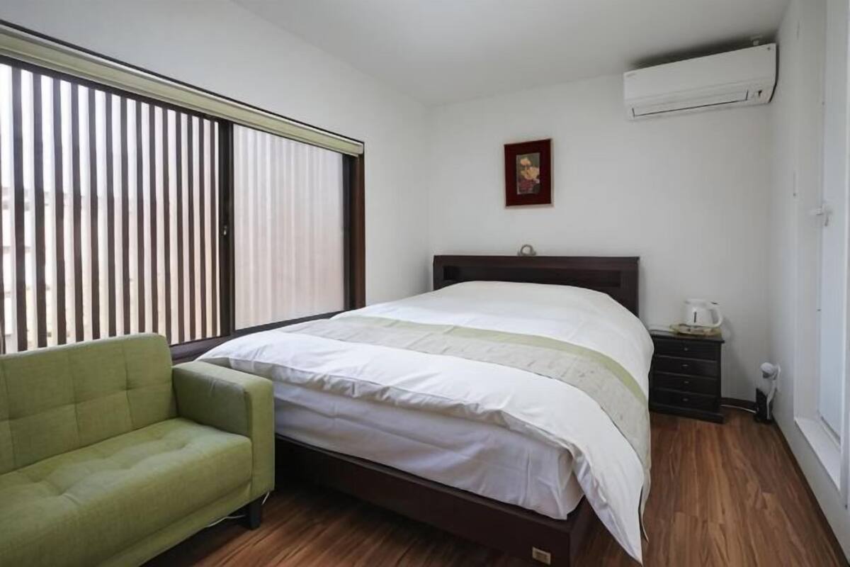 Quarto com cama, mesa de cabeceira, cadeira verde, janela com persianas e unidade de ar condicionado.