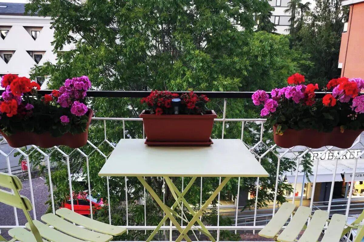 Uma varanda com duas cadeiras amarelas, uma mesa pequena e dois vasos de flores pendentes com flores vermelhas e roxas.