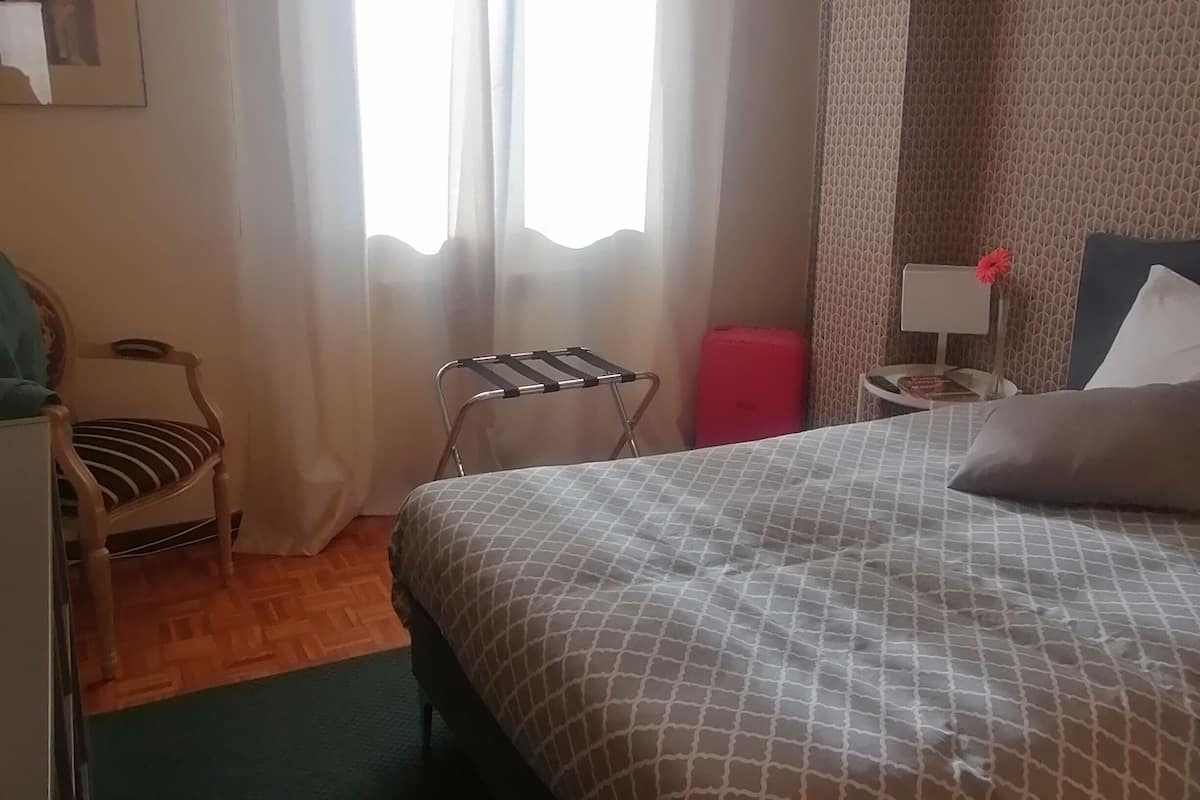 Quarto de hotel com cama, cadeira, mesa pequena e janela com cortinas.