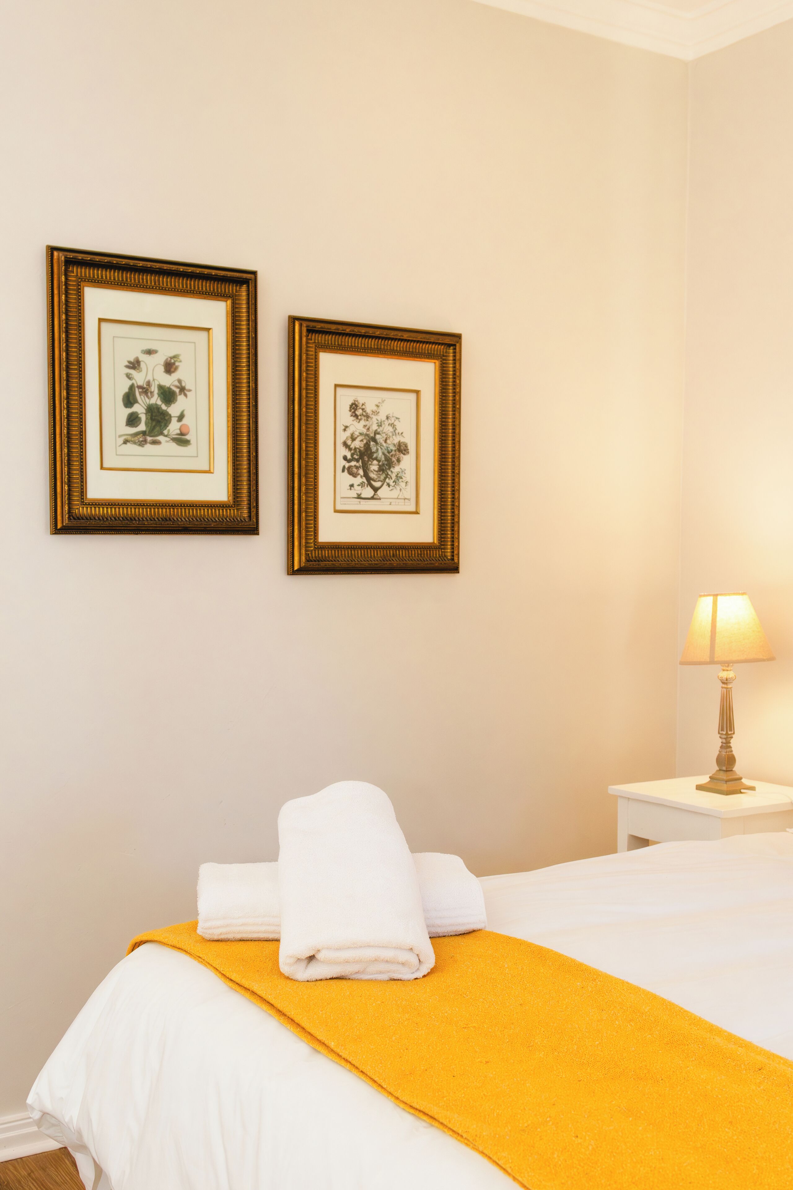 Quarto de hotel com cama, mesa de cabeceira com abajur e duas gravuras de plantas emolduradas na parede.
