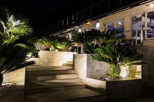 Área externa bem iluminada com degraus que levam a um edifício, cercada por grandes vasos de plantas e uma parede de pedra.