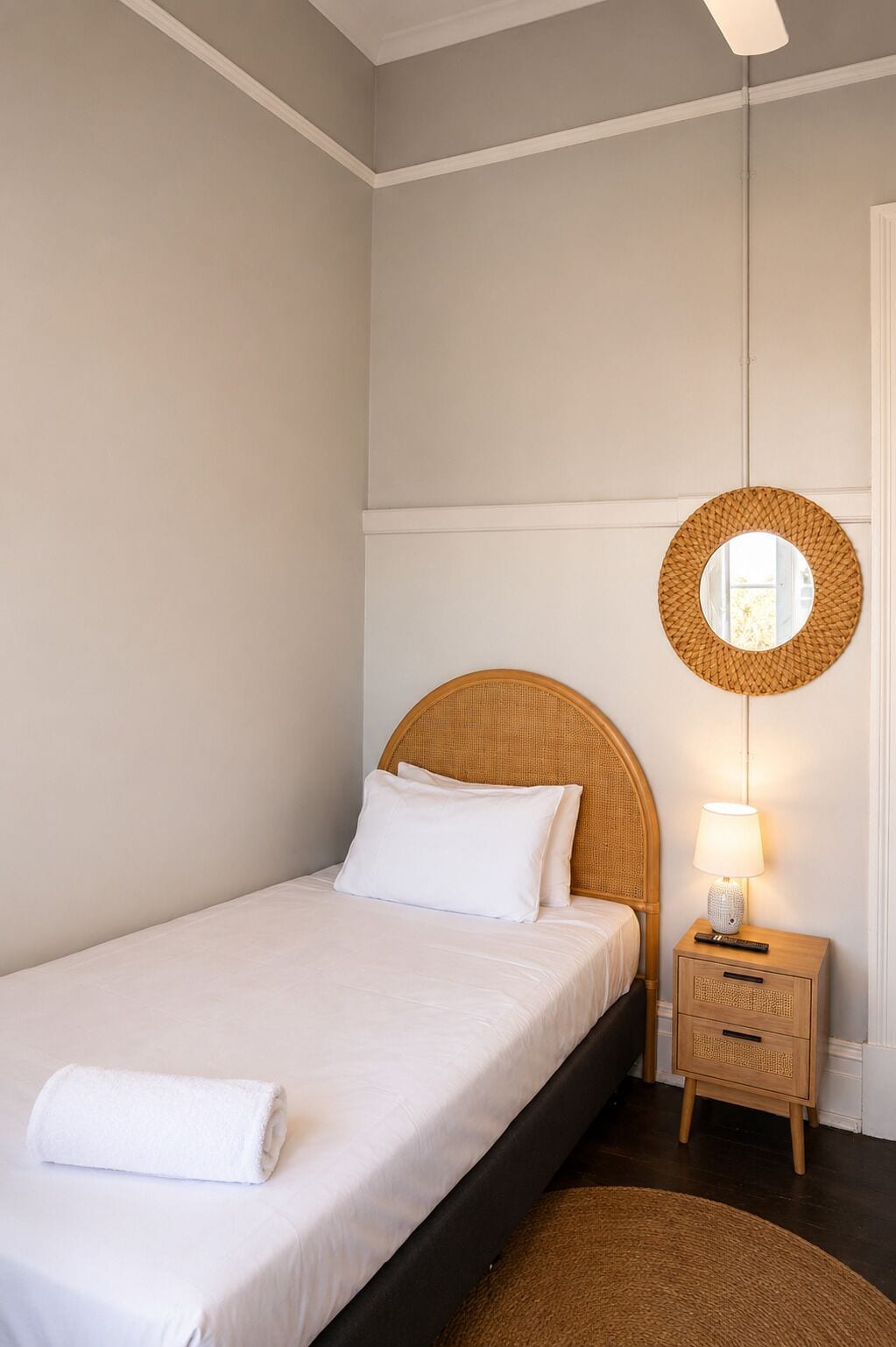 Uma cama bem arrumada com roupa de cama branca e uma toalha enrolada, um abajur, uma pequena mesa de cabeceira de madeira e um pendurar de parede em tecido.