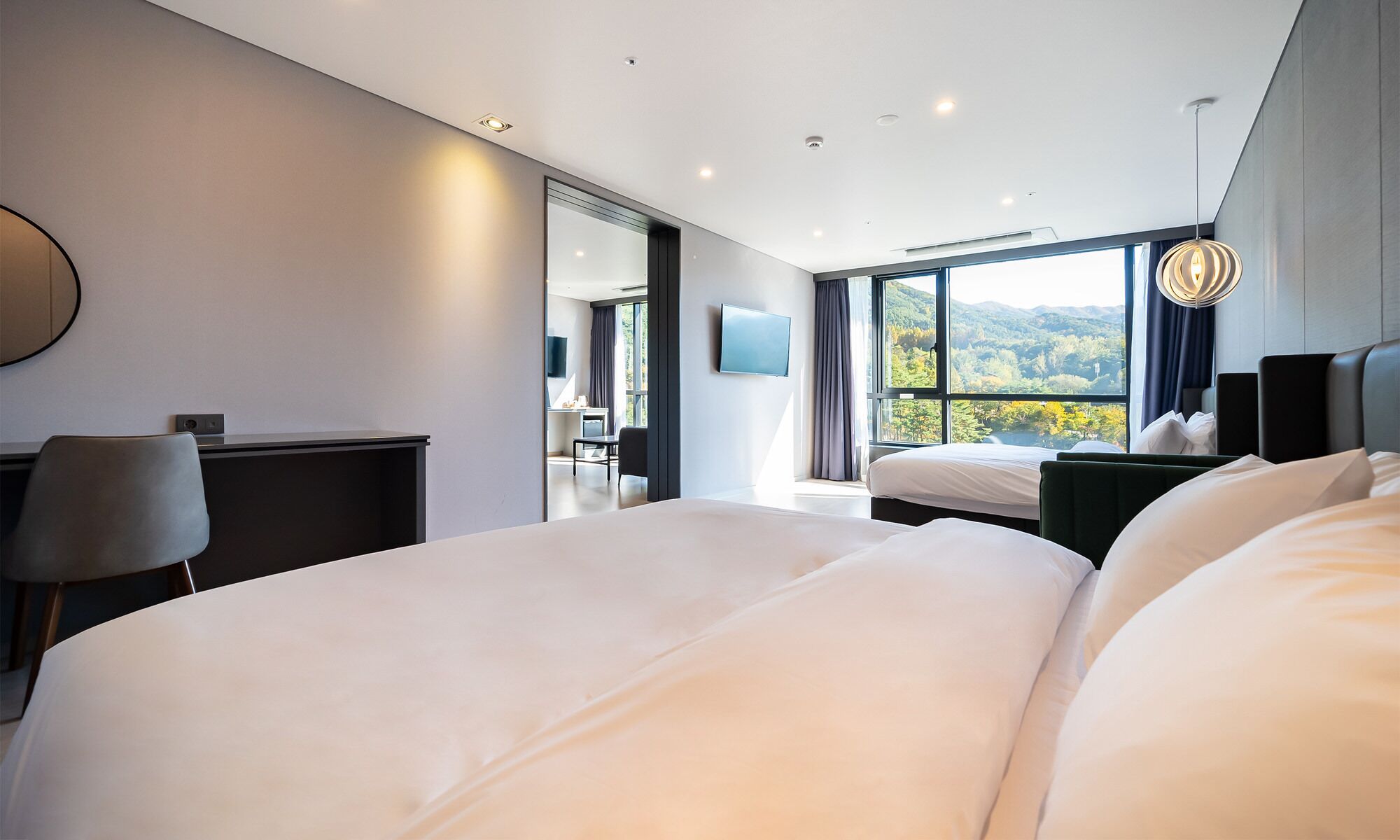 Quarto de hotel moderno com uma cama grande, uma escrivaninha e uma televisão. A janela oferece vista para uma paisagem montanhosa.